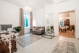 Uma sala de estar espaçosa com um sofá cinzento, uma mesa de centro de vidro e tapetes estampados. Um lustre pendurado no teto e uma grande janela garante iluminação natural.
