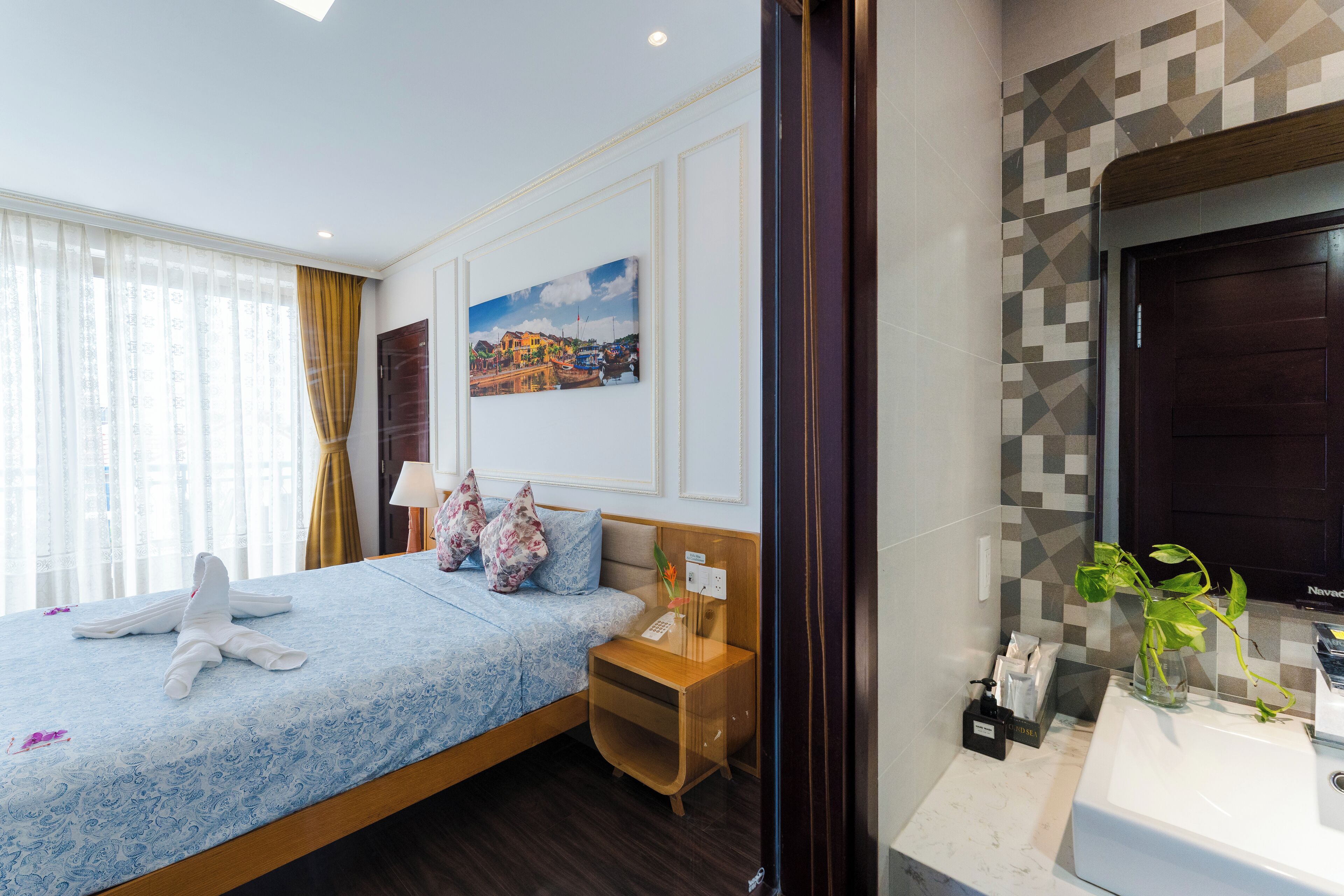Quarto de hotel com cama, mesa de cabeceira, pintura, banheiro com pia e espelho, e janela com cortinas.