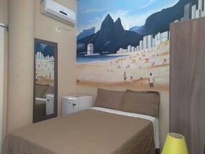 Quarto de hotel com cama, mesa de cabeceira, luminária, ar-condicionado fixado na parede e um espelho, além de um grande mural com tema de praia na parede.