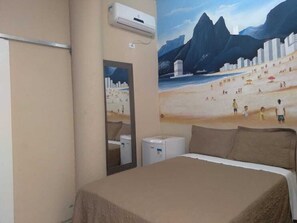 Quarto de hotel com cama, ar condicionado fixado na parede, espelho e pintura de uma praia.