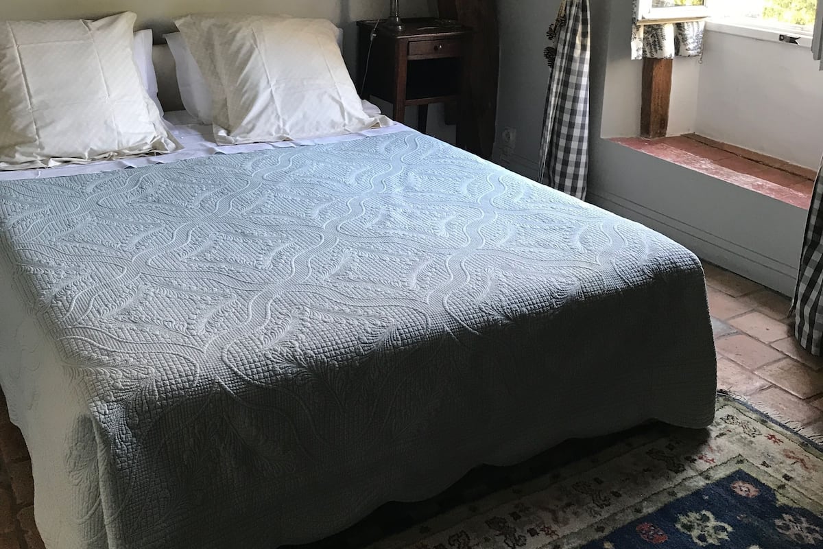 Um quarto com uma cama, uma mesa de cabeceira, uma luminária, um quadro na parede e uma janela com cortinas.