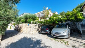 Uma rua residencial com dois carros estacionados, muros de pedra e uma casa com varanda.