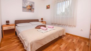 Quarto bem organizado com cama, mesa de cabeceira, gaveta e janela com cortinas leves.