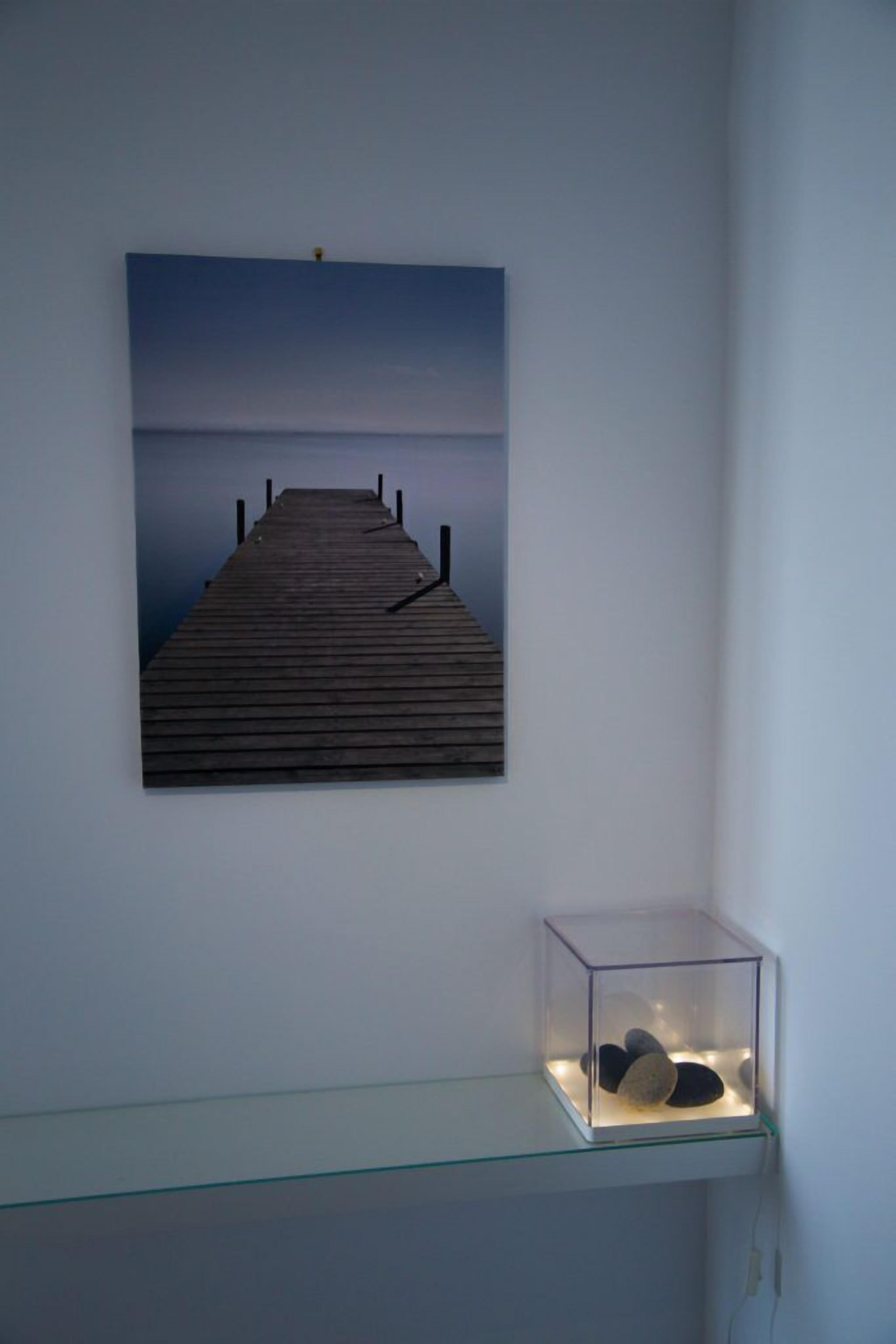 Um quadro de um cais numa parede, uma prateleira de vidro com um cubo cheio de pedras, e uma parede branca.
