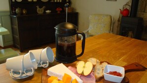Uma mesa de madeira com um tabuleiro de queijos, maçãs, pão e uma cafeteira.