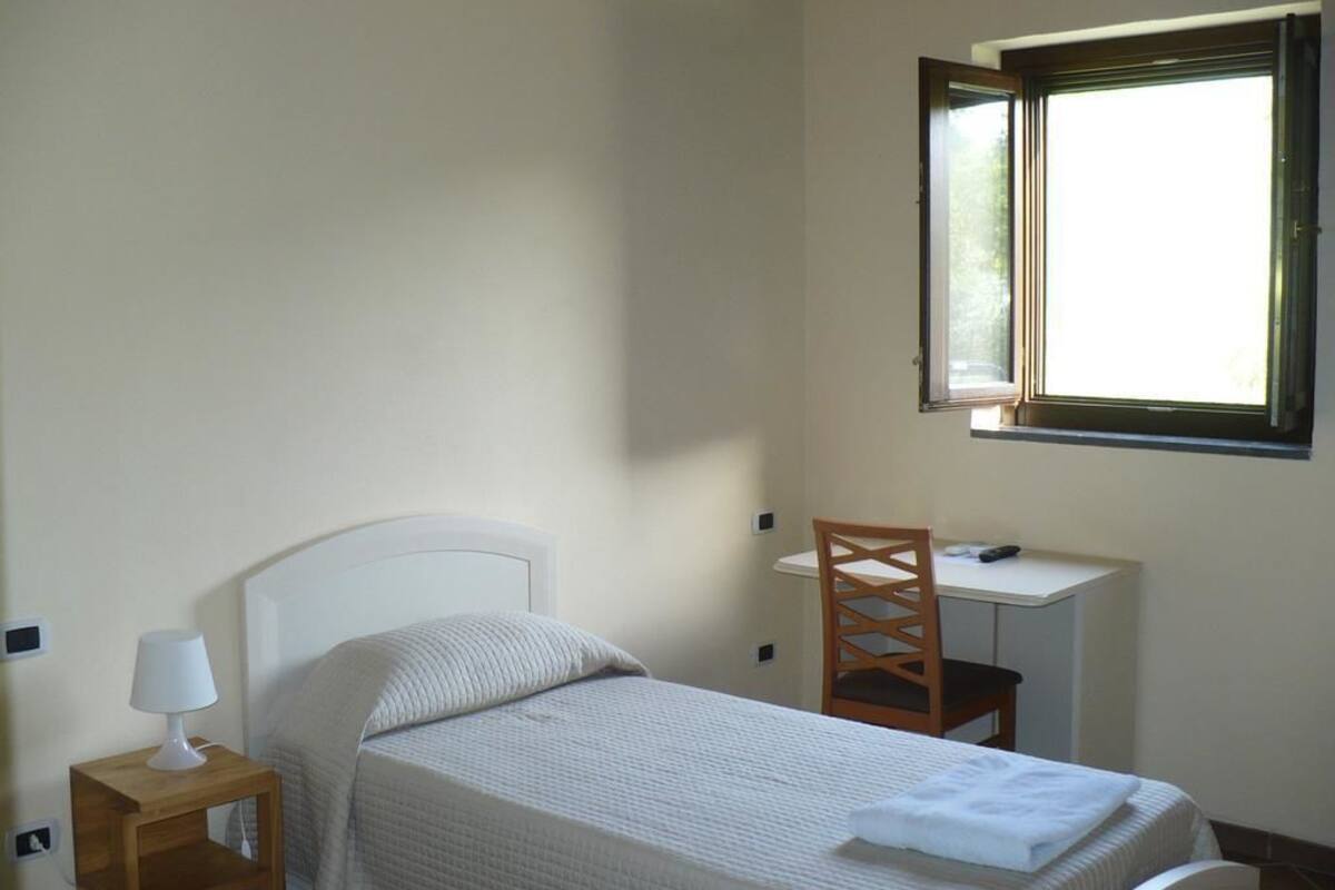 Quarto com cama, mesa de cabeceira com abajur, escrivaninha e uma cadeira.
