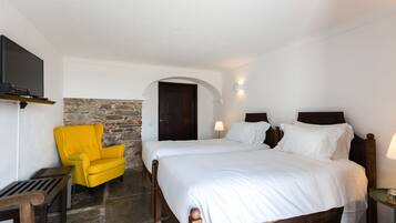 Quarto com duas camas, parede de pedra, porta de madeira, poltrona amarela e televisão de tela plana em uma estante.