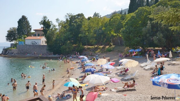 Uma praia com pessoas tomando sol e nadando, guarda-sóis e um edifício com parede de pedra.