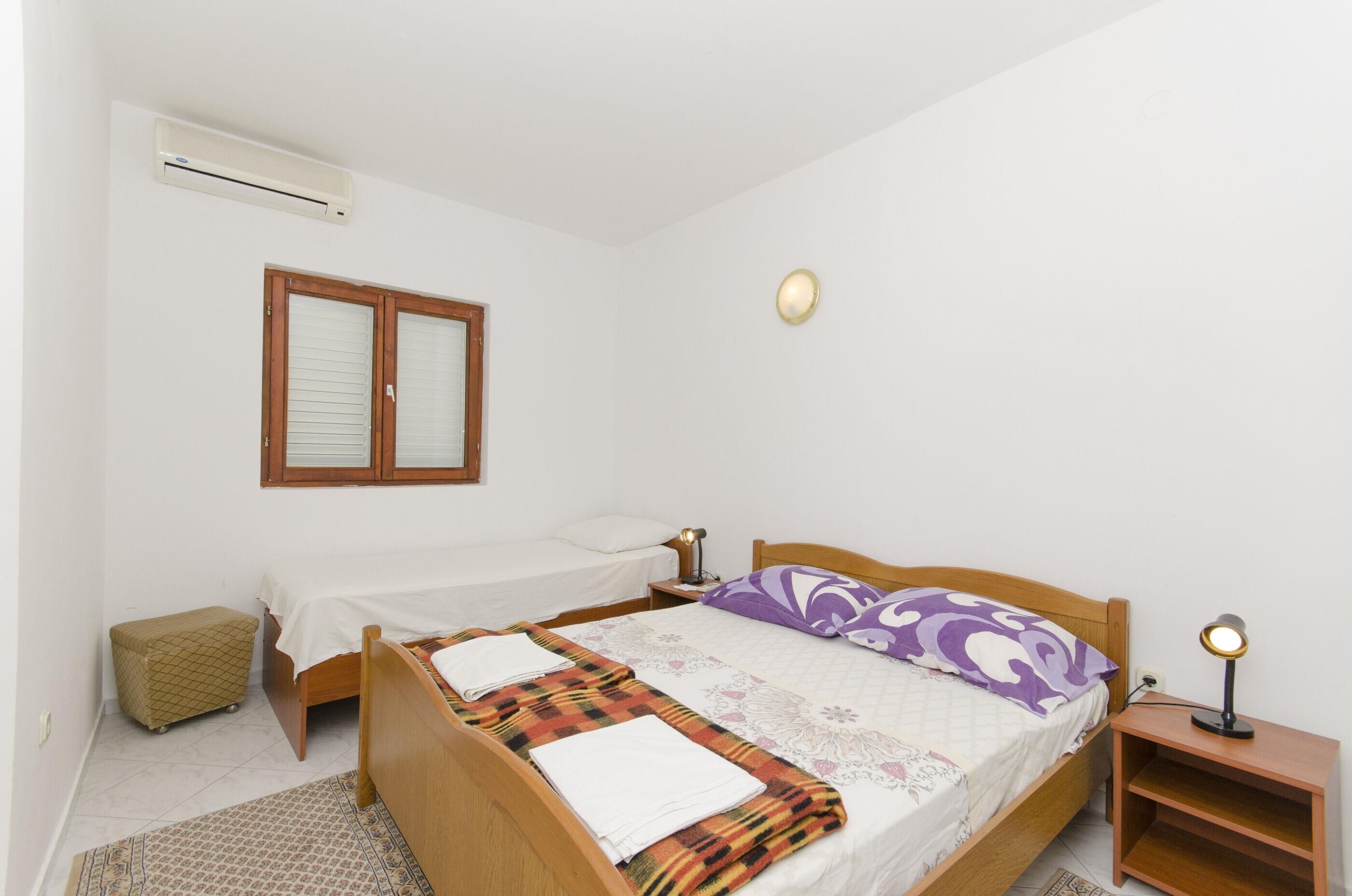 Quarto com uma cama de solteiro, mesa de cabeceira com abajur, pequena mesa de cabeceira com abajur, janela com persianas e unidade de ar condicionado na parede.