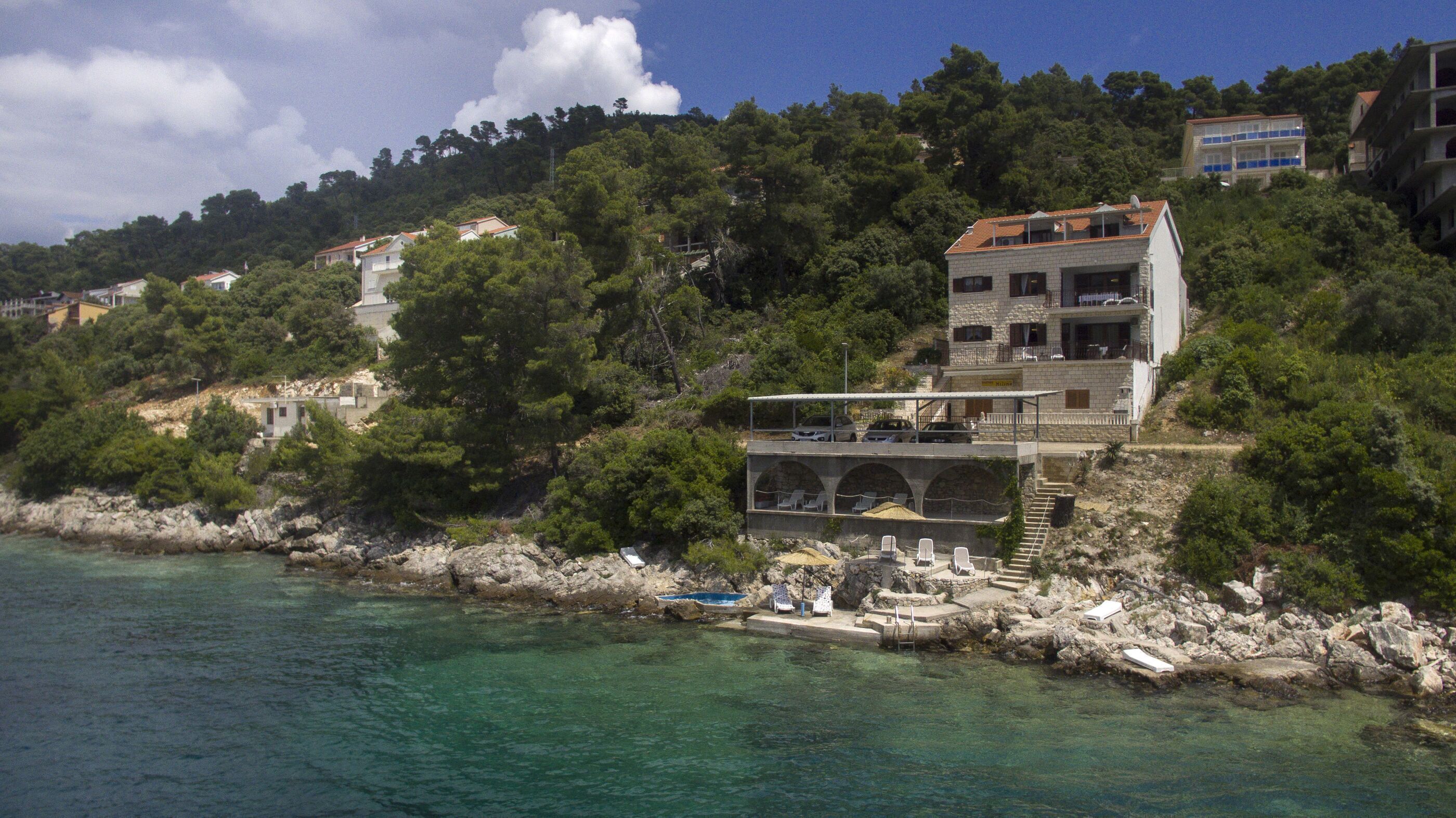 Um edifício costeiro com fachada de pedra, varanda com cadeiras e vista clara do mar azul-turquesa.