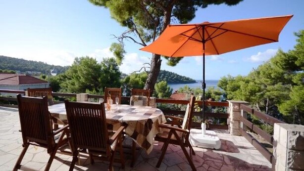 Um pátio externo com mesa e cadeiras, um guarda-sol laranja e uma vista panorâmica de um lago e colinas.