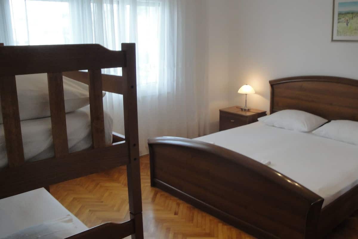 Uma cama de madeira com roupa de cama branca em um quarto com piso de madeira e uma mesinha de cabeceira com abajur.