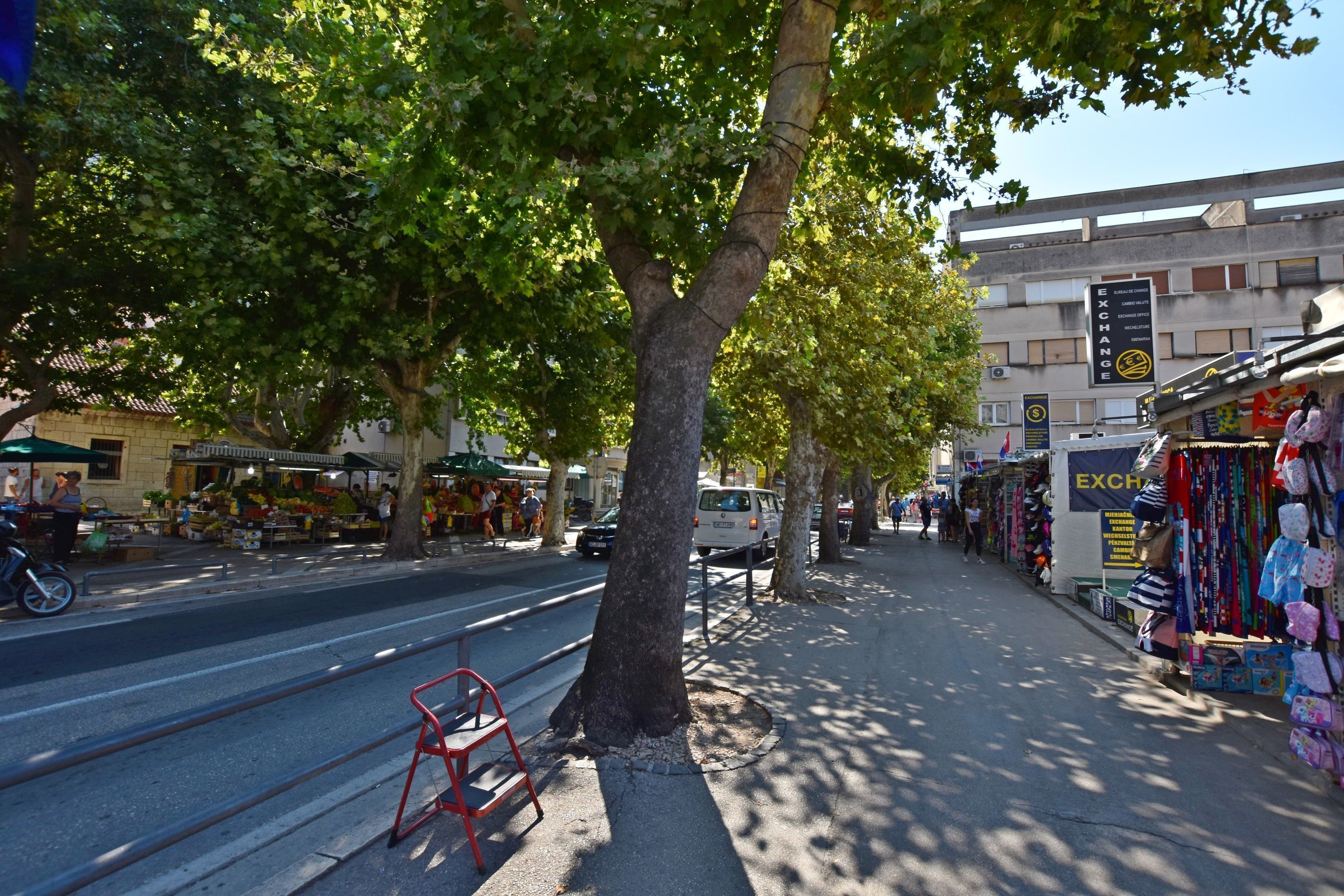 Rua com linha de bonde, árvores e uma banca de mercado vendendo bolsas coloridas.