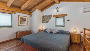 Quarto com teto de madeira, uma cama grande com colcha acolchoada, duas mesas de cabeceira com luminárias, uma parede de destaque em pedra e uma janela com cortina.