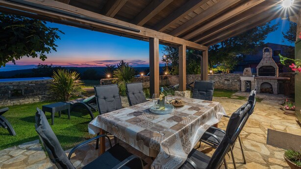 Um pátio externo com uma mesa posta para refeições, rodeado por cadeiras e uma lareira de pedra ao fundo.