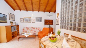 Uma sala de estar com teto de madeira, um sofá floral, uma poltrona laranja, uma mesa de jantar posta com uma fruteira e vista através de um muro de vidro.