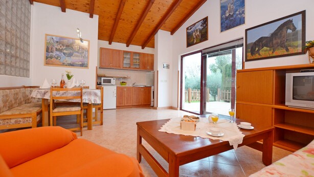 Uma sala de estar acolhedora com teto de madeira, um espaço de jantar com mesa posta para dois e uma cozinha ao fundo.