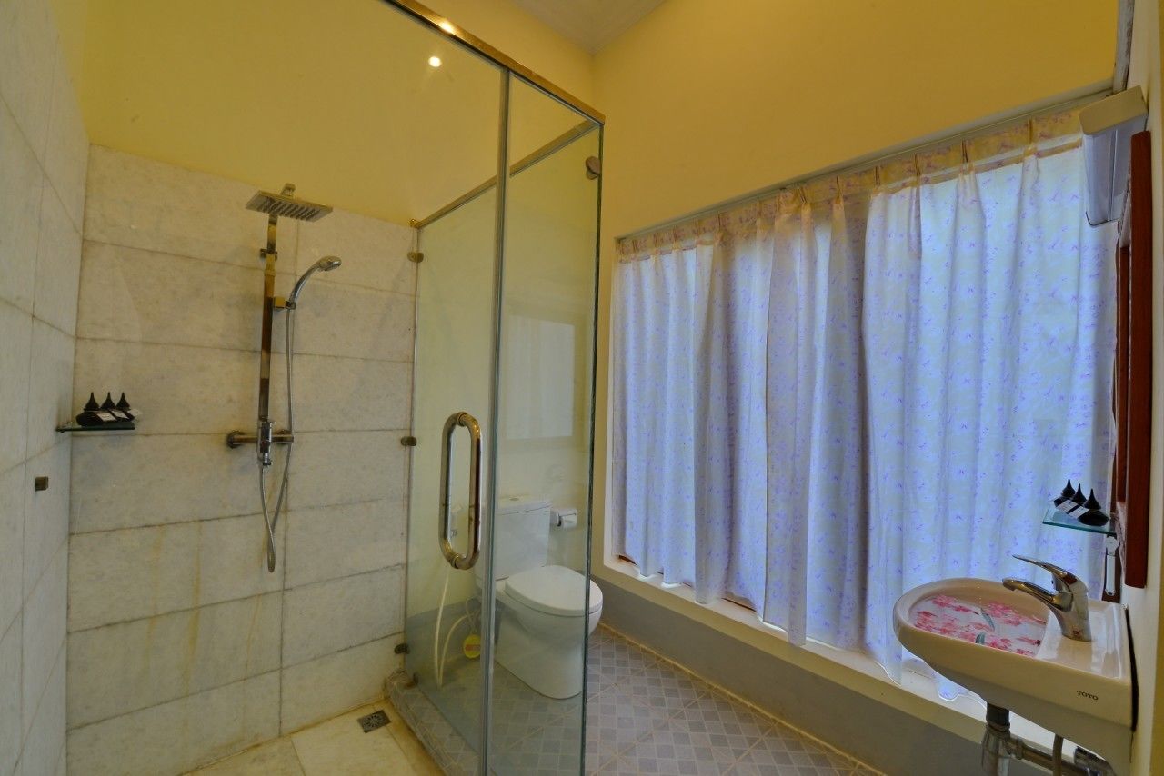 Casa de banho com chuveiro de vidro, sanita, lavatório com espelho e janela com cortinas.
