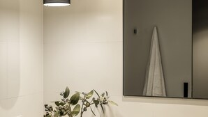 Casa de banho com parede revestida a azulejos brancos, espelho, lavatório com torneira, uma planta e uma vela.