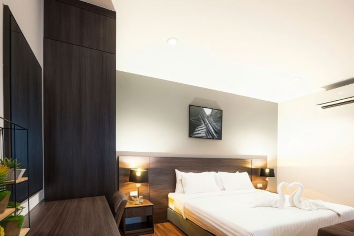 Quarto de hotel moderno com uma cama grande, duas mesas de cabeceira com abajures, uma cabeceira em madeira escura, uma obra de arte emoldurada na parede e uma planta no lado esquerdo.