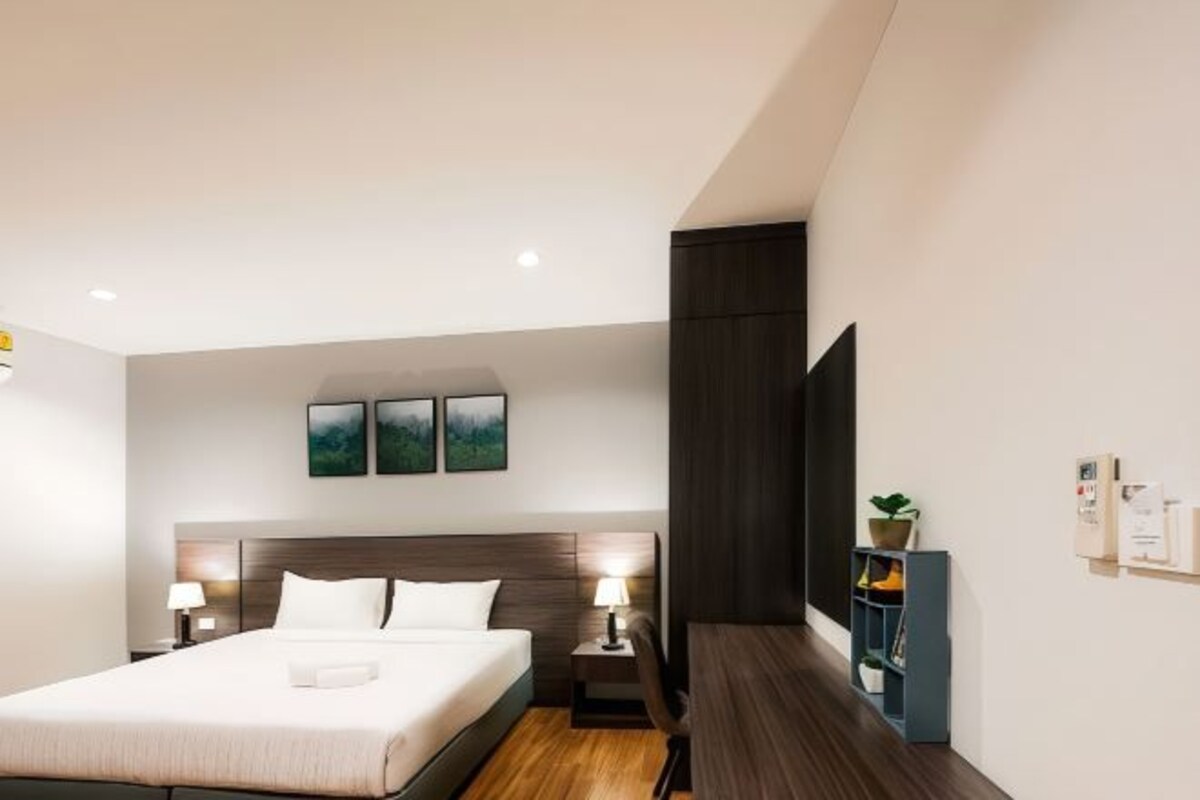 Quarto de hotel moderno com uma cama grande, abajures, cabeceira em madeira escura e uma pequena escrivaninha com cadeira. Três quadros estão pendurados na parede acima da cama.