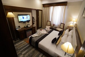 Quarto de hotel com uma cama grande, uma escrivaninha com televisão, uma cadeira e um abajur.