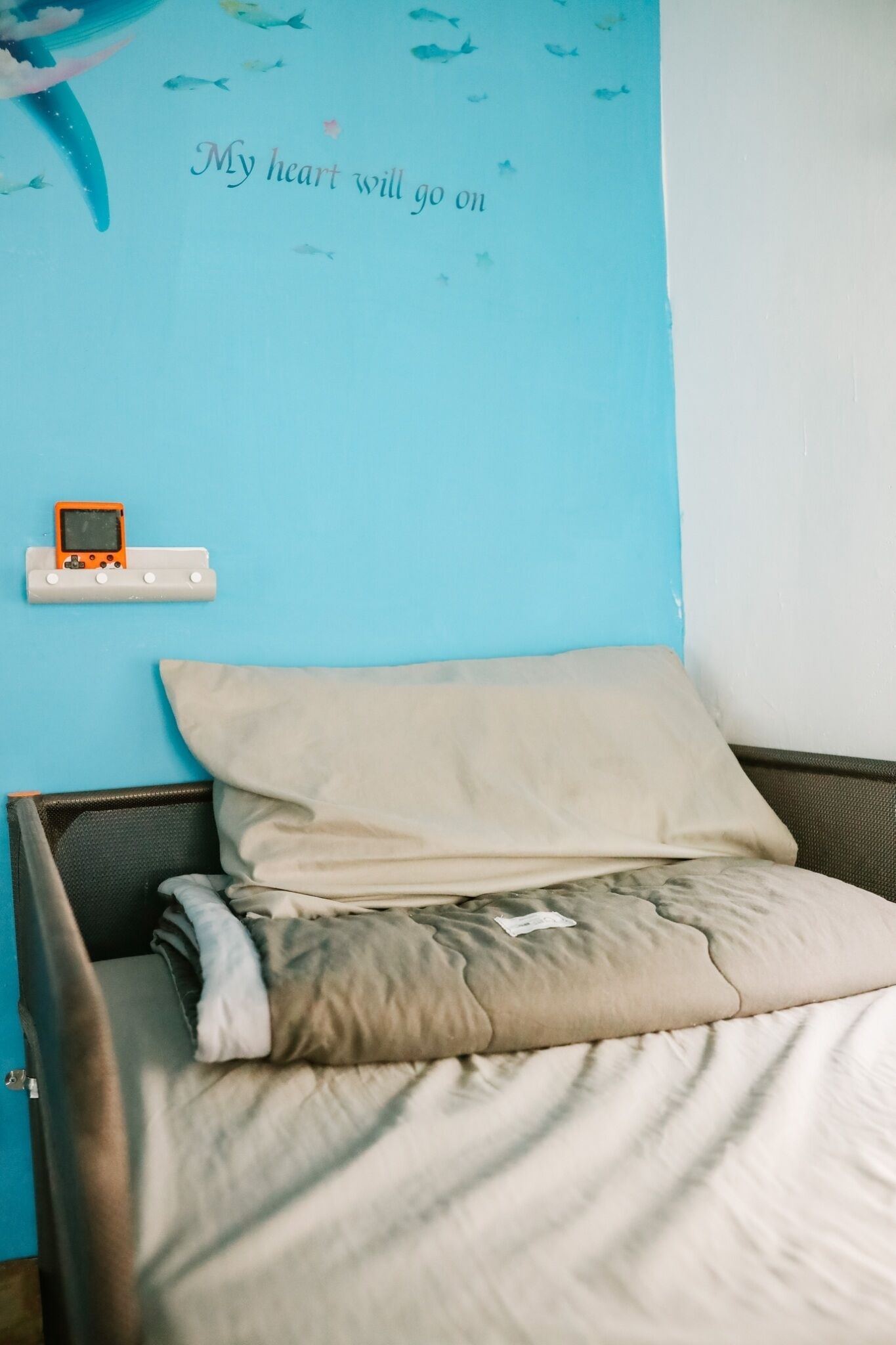 Cama bem arrumada com roupa de cama bege, prateleira na parede com um pequeno aparelho laranja e um adesivo de parede com o texto "My heart will go on" e ilustrações de peixes.