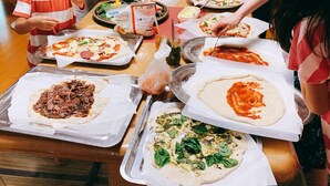 Uma família preparando pizzas caseiras em uma cozinha com micro-ondas e diversos utensílios de cozinha.