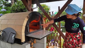 Forno a lenha de tijolos ao ar livre, com fogo no interior, uma pá de pizza e uma cesta de toras.
