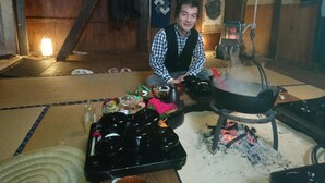 Uma pessoa sentada em um quarto tradicional com piso de tatame, um forno a carvão e uma grande panela preta sobre um suporte.