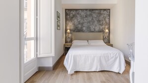 Um quarto com uma cama grande, duas mesas de cabeceira com luminárias, uma arte abstrata na parede em tons de cinza e uma janela com persianas.
