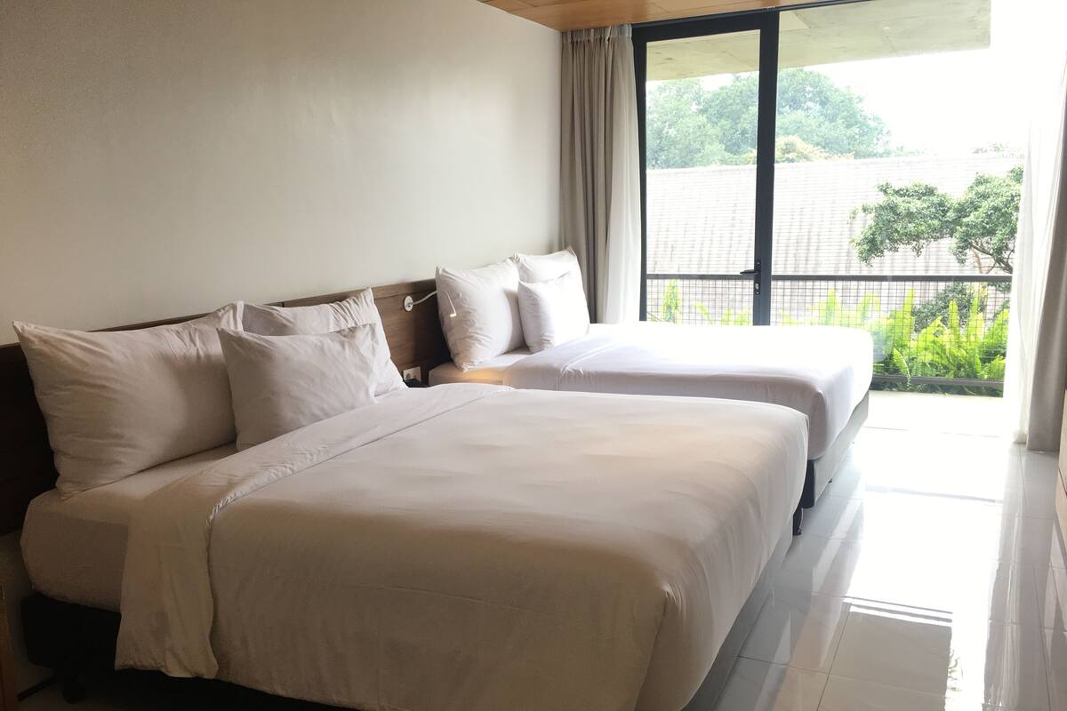 Quarto de hotel com duas camas, uma janela grande e vista para o verde lá fora.