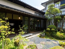Um edifício japonês tradicional com entrada de vidro moderna, rodeado por um jardim bem cuidado com pedras, musgo e diversas plantas.