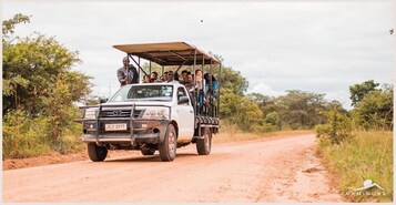 Uma caminhonete branca com rack de teto transportando passageiros em um caminho de terra, cercada por vegetação.