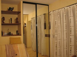 Quarto com cama, espelho, prateleiras com objetos decorativos e cortina de chuveiro estampada.