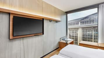 Quarto de hotel moderno com uma televisão grande fixada na parede, uma pequena mesa com tampo de vidro e uma janela ampla com vista para a cidade.
