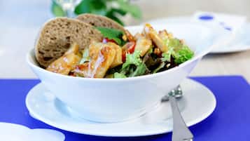 Uma saladeira branca com salada, croutons e uma colher sobre uma toalha de mesa azul.