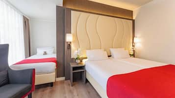 Quarto de hotel com duas camas, uma cadeira, mesa de cabeceira com abajur e um vaso decorativo.
