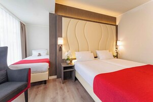Quarto de hotel com duas camas, uma cadeira, mesa de cabeceira com abajur e um vaso decorativo.