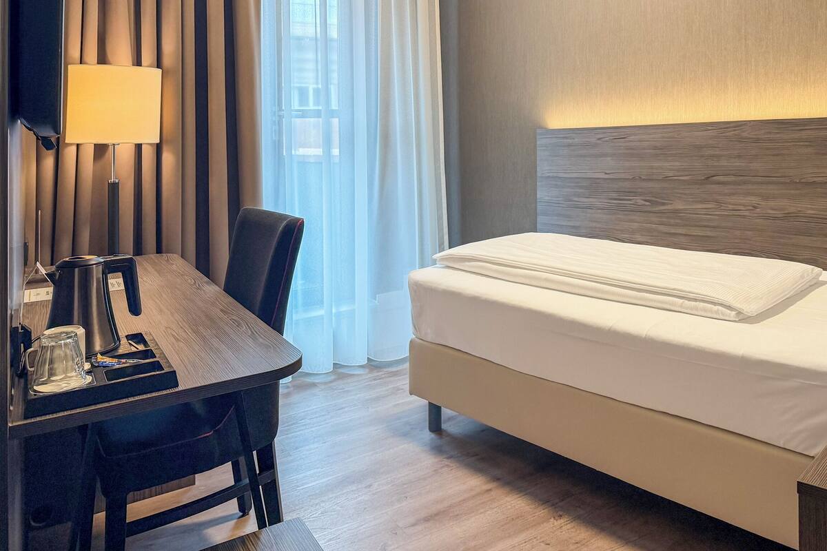 Quarto de hotel com cama, escrivaninha com luminária, cadeira e janela com cortinas.