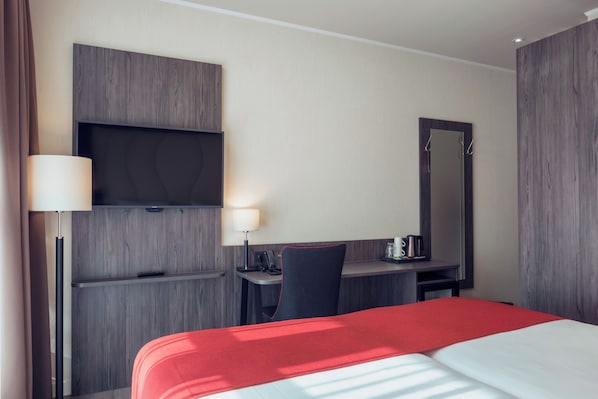 Quarto de hotel com cama, escrivaninha, cadeira, televisão, abajur e espelho.