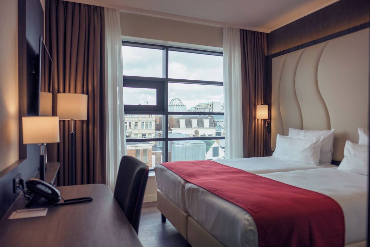 Quarto de hotel com uma cama grande, uma mesa de trabalho com telefone e vista da cidade através de amplas janelas.