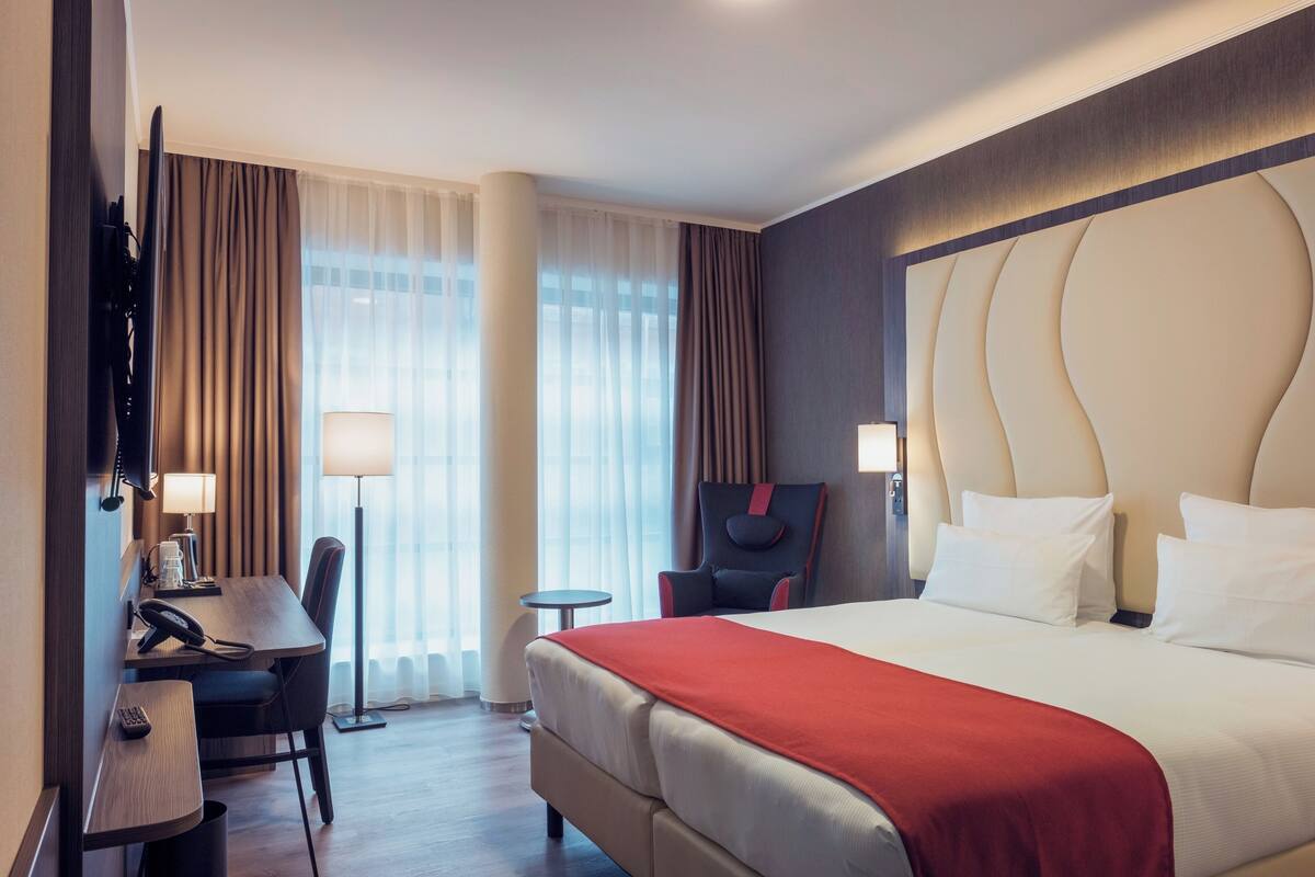 Quarto de hotel com uma cama, escrivaninha, cadeira, luminária e televisão.