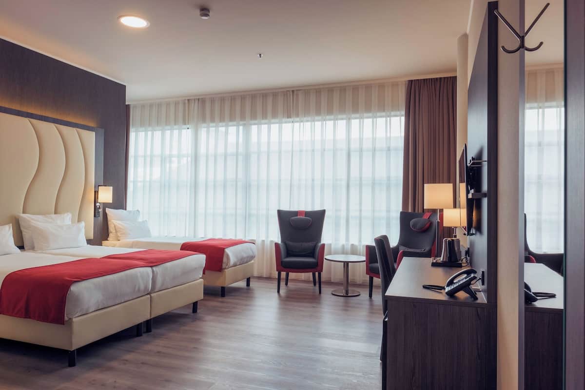 Quarto de hotel com cama, duas poltronas, uma mesa pequena, escrivaninha, telefone e espelho.