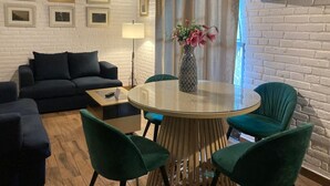 Uma sala de estar acolhedora com uma mesa de vidro redonda, cadeiras verdes, um sofá preto e uma TV fixada na parede.