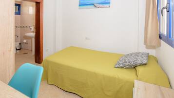 Quarto pequeno com uma cama de solteiro, colcha amarela, travesseiro, cadeira azul, janela com cortinas e um quadro de uma cena de praia na parede.