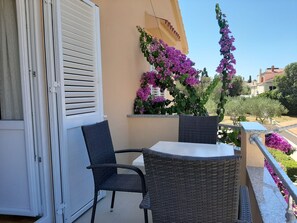 Varanda com cadeiras de vime e uma mesa, janela com persianas e flores roxas.