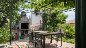 Um pátio externo com mesa e cadeiras, rodeado por plantas em vasos e um forno de tijolos.