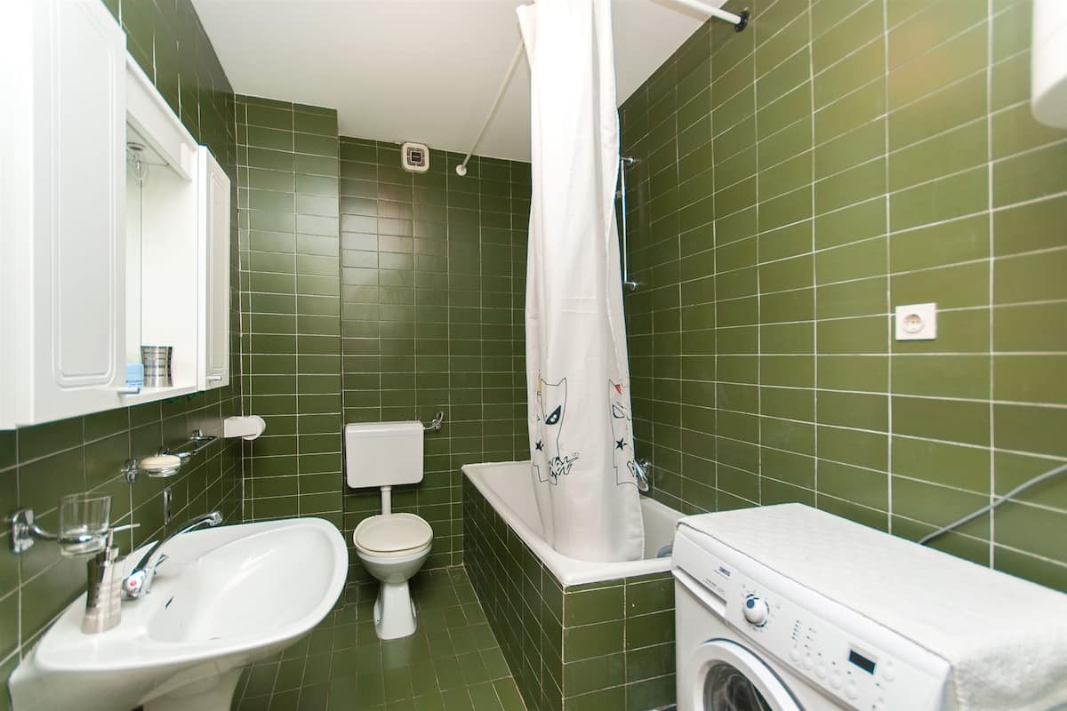 Casa de banho com azulejos verdes, pia branca, sanita, banheira e máquina de lavar roupa.