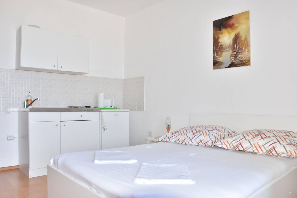Quarto com cama, mesa de cabeceira, quadro na parede e um espaço com cozinha compacta.
